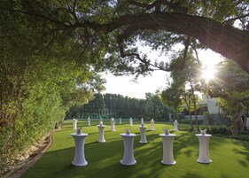 Um jardim com mesas brancas e plantas em vasos, rodeado por árvores e um caminho.