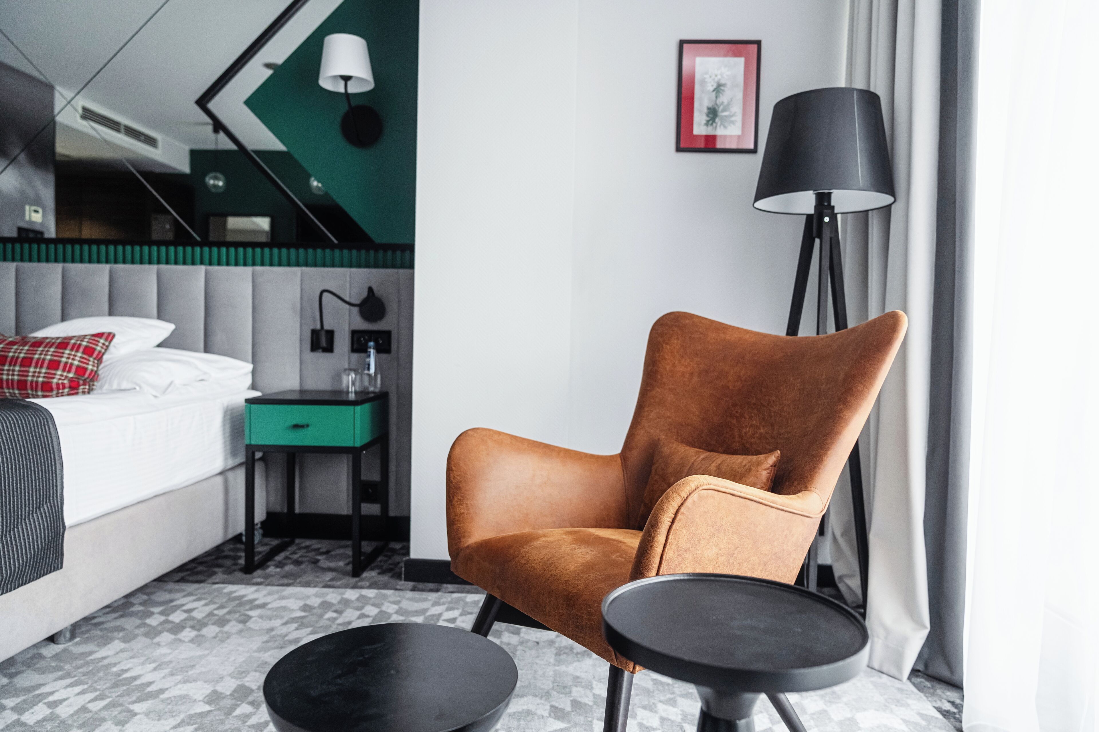 Quarto de hotel moderno com uma cama, uma mesa de cabeceira verde, uma poltrona de couro marrom, um abajur de chão preto e uma pequena mesa redonda.