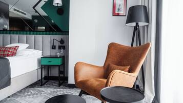Quarto de hotel moderno com uma cama, uma mesa de cabeceira verde, uma poltrona de couro marrom, um abajur de chão preto e uma pequena mesa redonda.