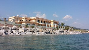 Um imóvel costeiro com uma construção grande e várias varandas, um litoral rochoso e vista clara do mar.