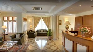 Um lobby amplo com uma mesa de recepção, área de estar com sofás e uma grande janela com cortinas.