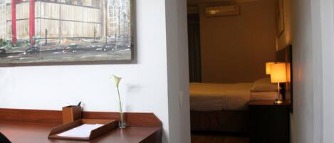 Quarto de hotel com escrivaninha de madeira, pintura emoldurada de uma vista urbana, mesa de cabeceira com abajur e cama com roupa de cama branca.