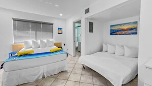 Quarto de hotel com cama, janela com persianas, pintura na parede e banheiro visível através de uma porta aberta.