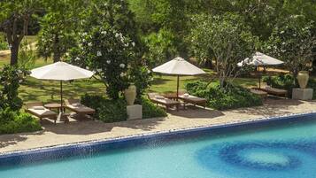 Uma área à beira da piscina com espreguiçadeiras e guarda-sóis, cercada por vegetação exuberante e uma piscina azul cristalina.