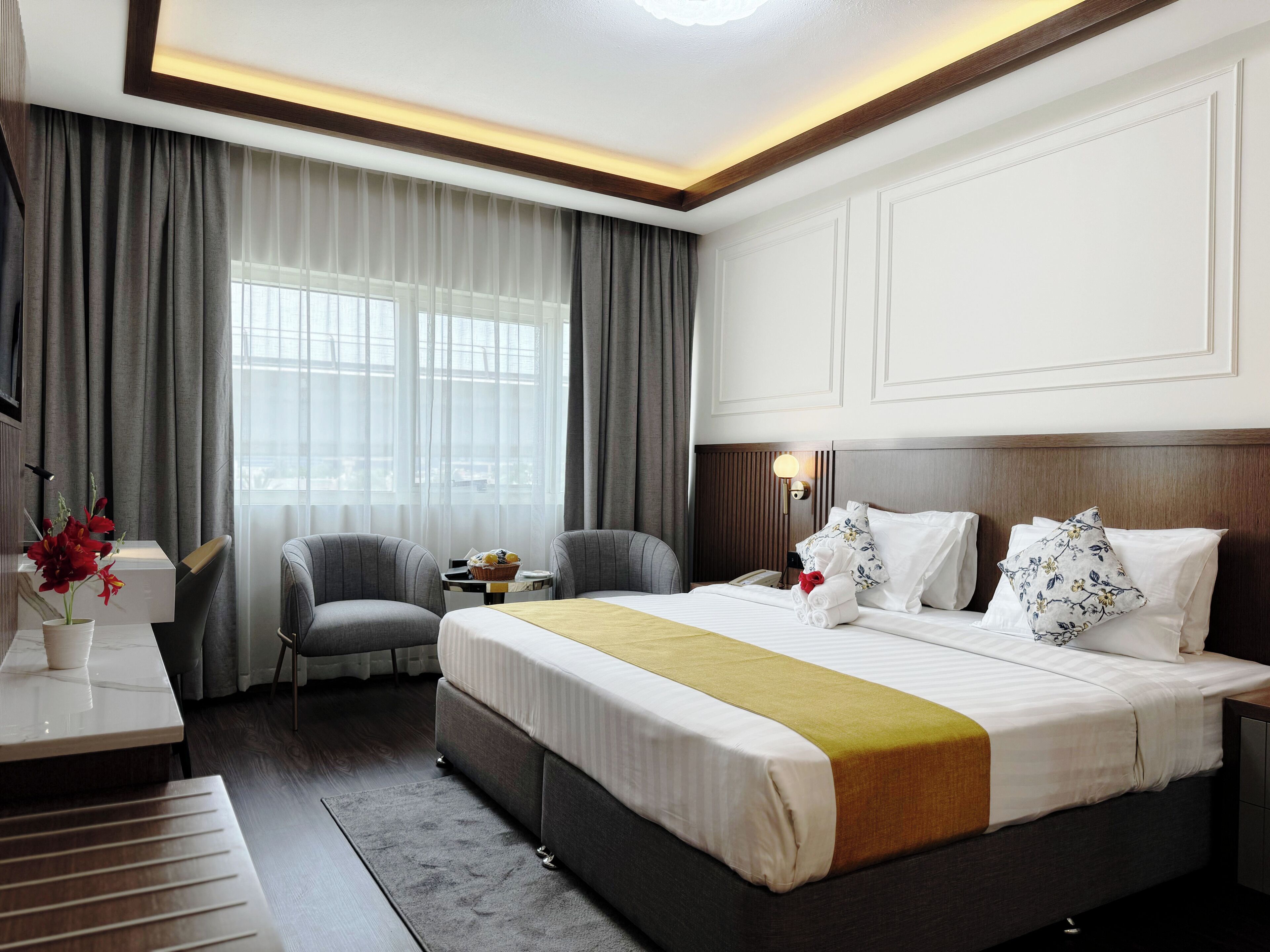 Quarto de hotel com uma cama grande, duas poltronas, uma mesinha e uma escrivaninha com abajur e flores.