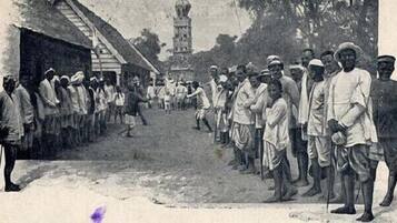 Cena histórica com pessoas vestindo roupas tradicionais, um edifício com escadaria e uma torre ao fundo.