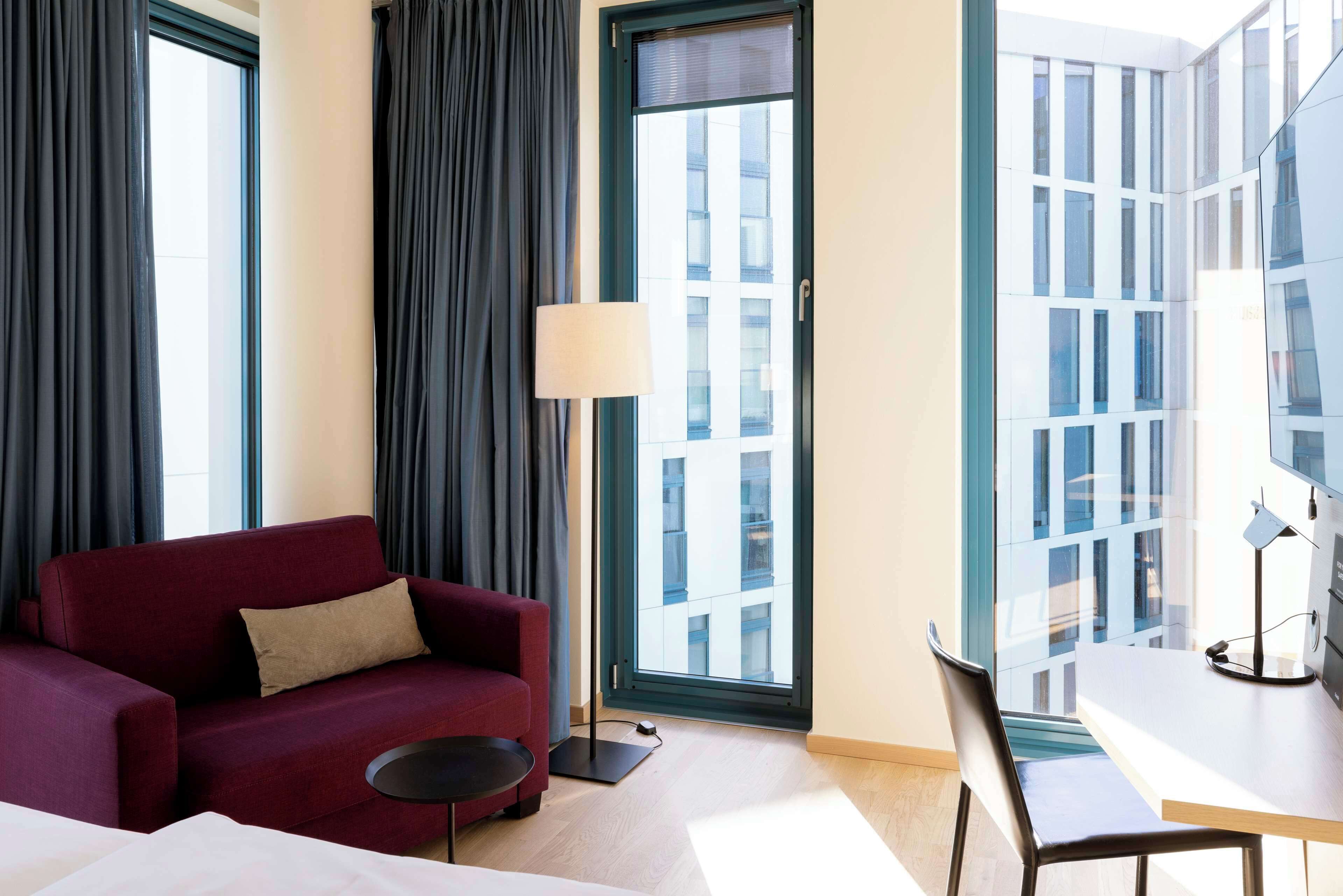 Quarto de hotel com sofá vermelho, uma mesa redonda pequena, escrivaninha com luminária e vista para um prédio através de uma janela grande.