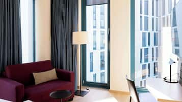 Quarto de hotel com sofá vermelho, uma mesa redonda pequena, escrivaninha com luminária e vista para um prédio através de uma janela grande.
