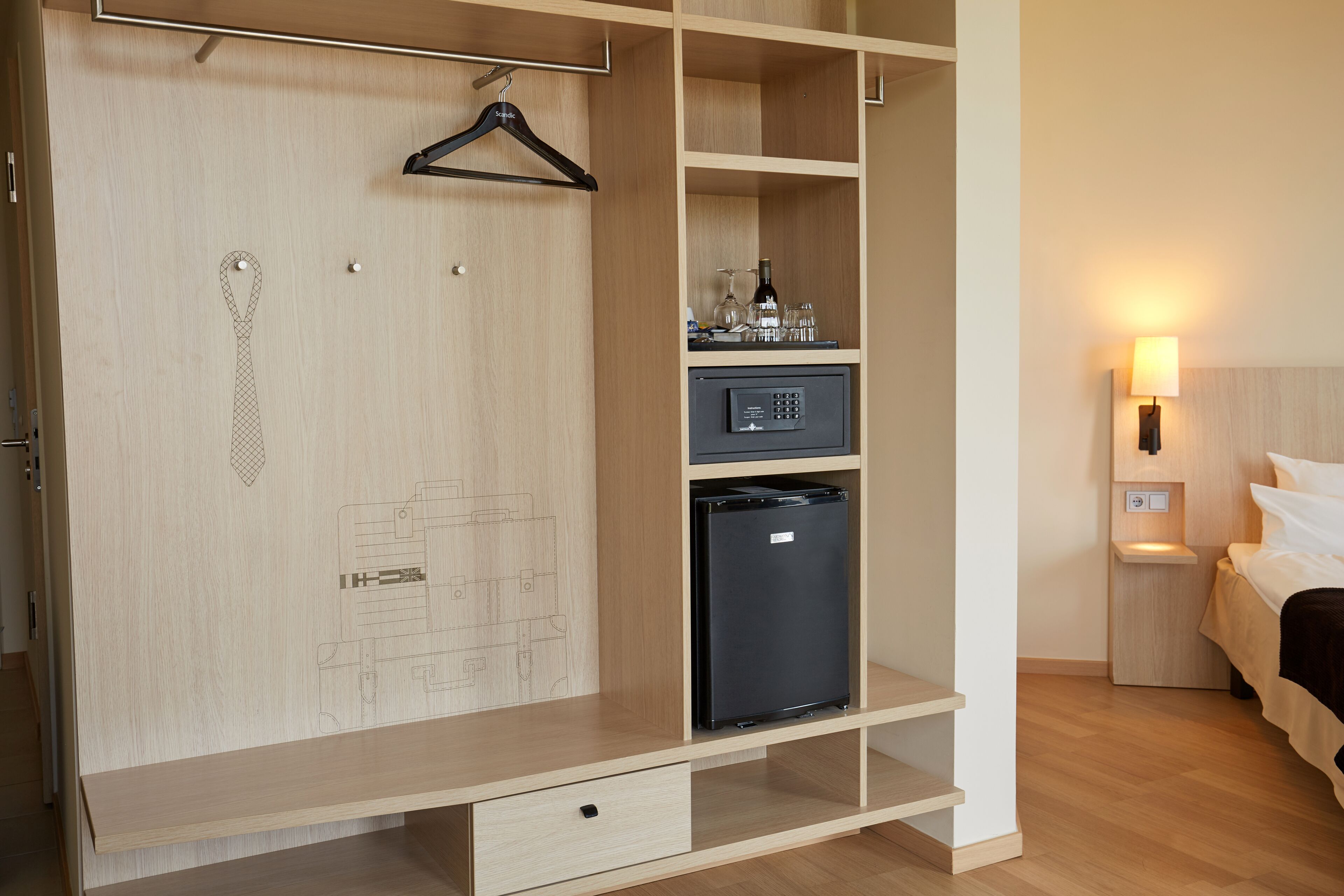 Quarto de hotel com guarda-roupa de madeira, pequeno frigorífico preto, mesa de cabeceira com candeeiro e cama com roupa de cama branca.