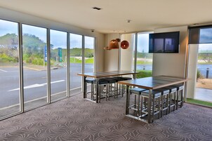 Um interior moderno com janelas amplas, uma mesa de madeira, cadeiras de metal e uma TV montada na parede.