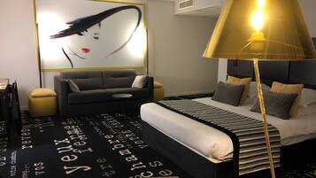 Quarto de hotel moderno com uma cama grande, um sofá, uma mesa redonda pequena e um abajur de chão.
