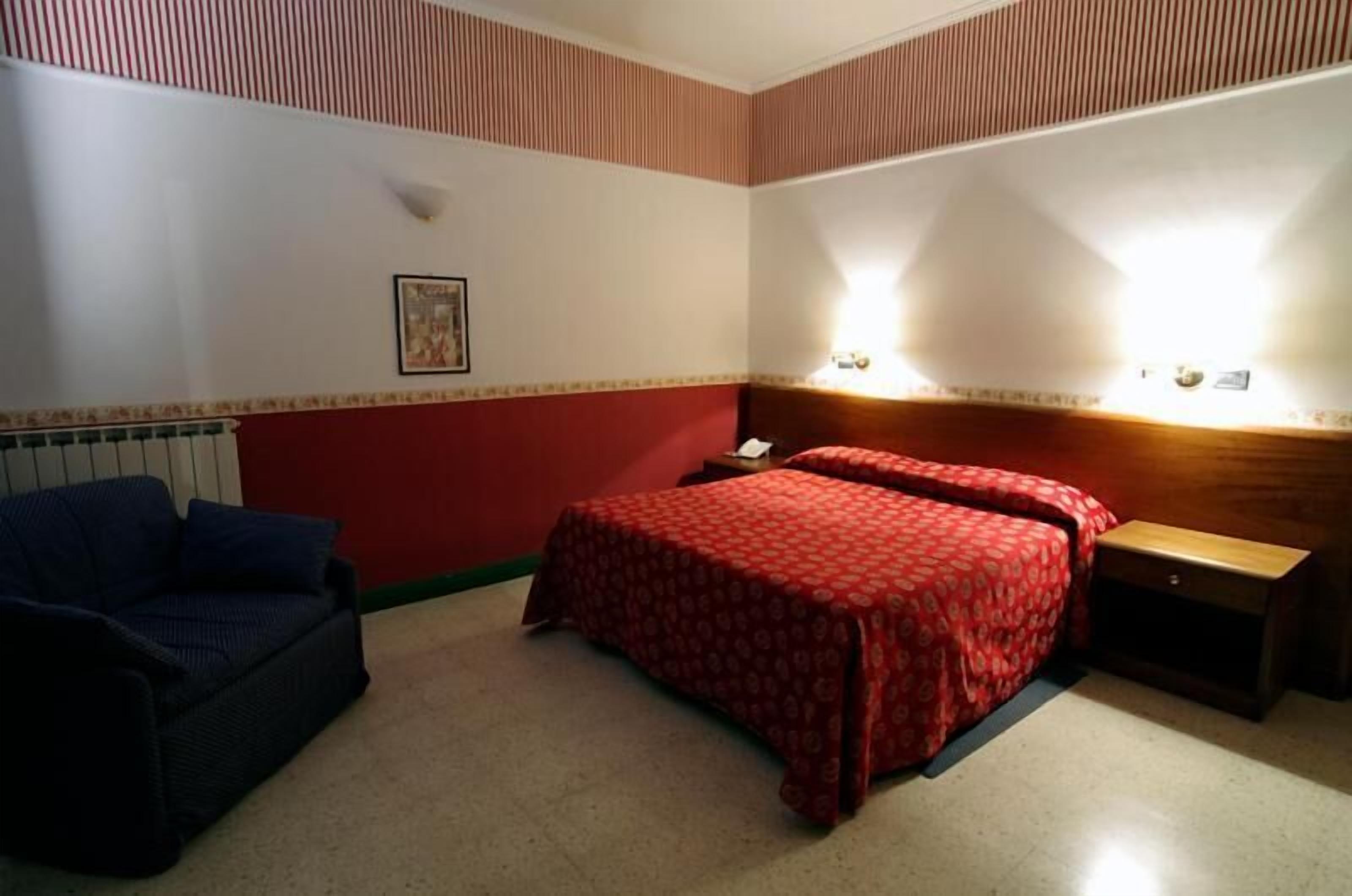 Quarto de hotel com cama, mesa de cabeceira, um banquinho de madeira, um sofá azul e um quadro na parede.