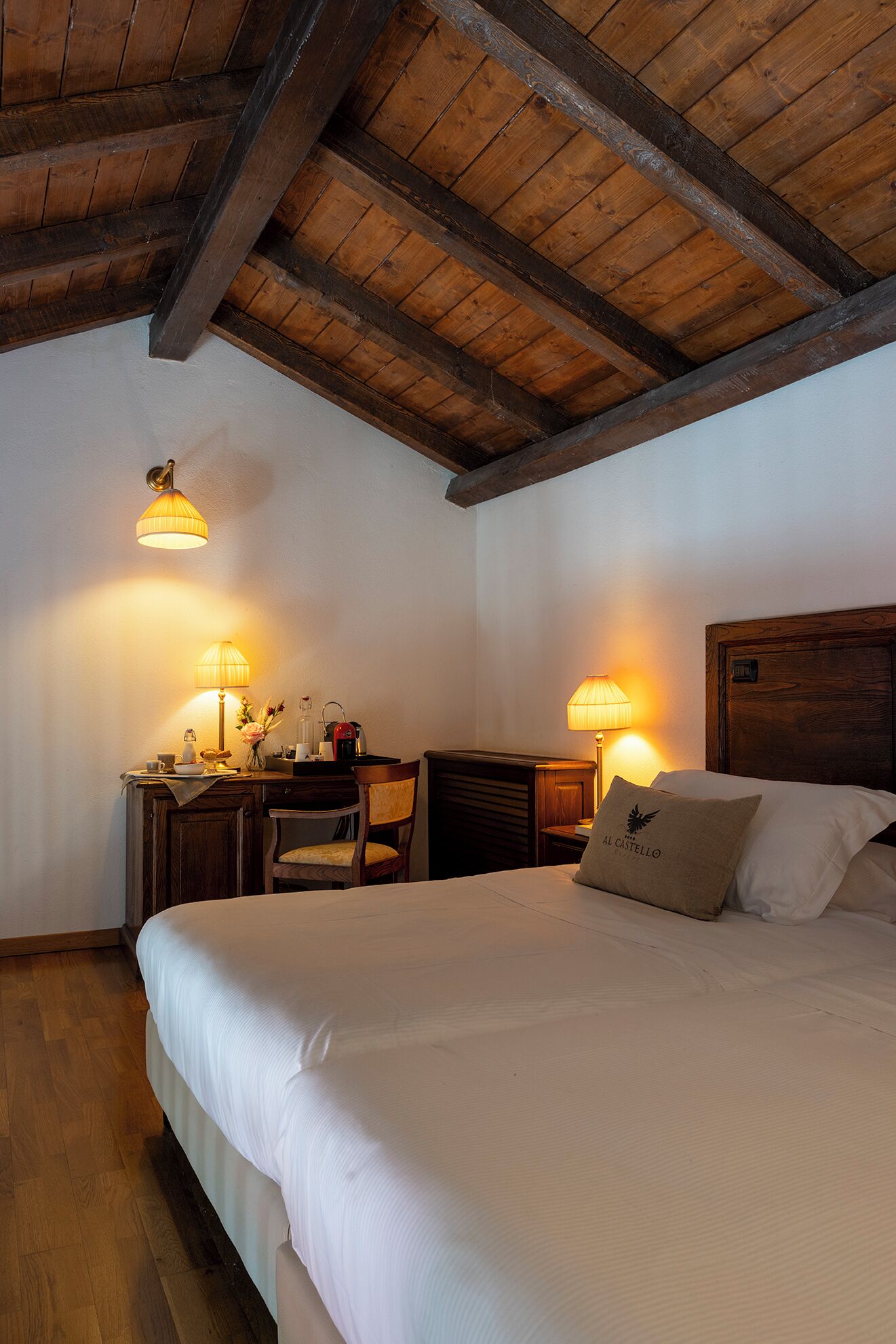 Quarto de hotel com teto de madeira, cama com roupa de cama branca, duas luminárias de cabeceira, escrivaninha com cadeira e mesa de cabeceira com abajur.