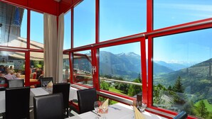 Restaurante com grandes janelas proporcionando vista para as montanhas. Mesas montadas com toalhas brancas e taças de vinho.