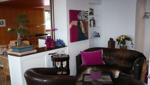 Sala de estar com sofá de couro marrom, mesa de centro e planta em vaso. Há uma mesa pequena com um vaso de flores e uma estante com objetos decorativos.