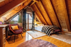 Um quarto de sótão acolhedor com teto de madeira, uma poltrona vermelha macia, uma cama com manta de pele e uma grande janela com cortinas.