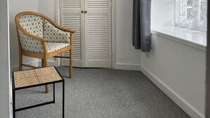 Quarto com carpete cinza, uma cadeira de madeira com almofada estampada, um guarda-roupa branco e uma janela com cortina cinza.