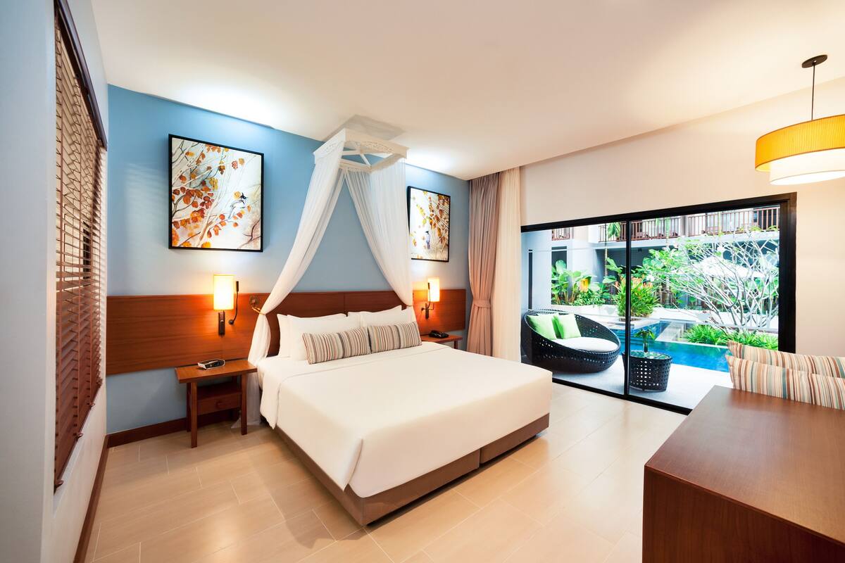Quarto de hotel com cama grande, mesas de cabeceira, escrivaninha, cadeira, janela ampla com vista para piscina e vegetação, e duas luminárias de parede.