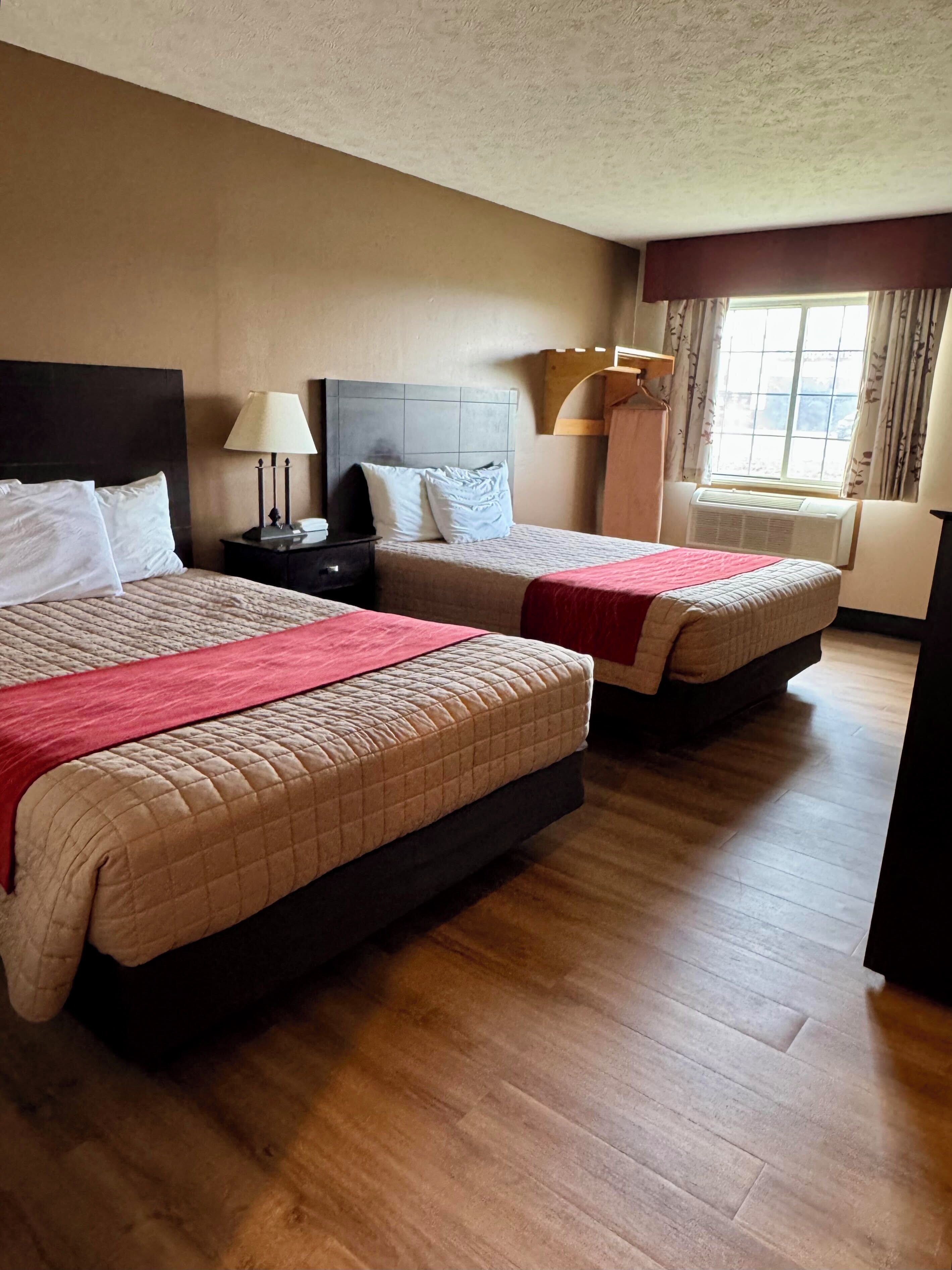 Quarto de hotel com duas camas, uma mesinha de cabeceira, uma luminária, uma janela com cortinas e piso de madeira.