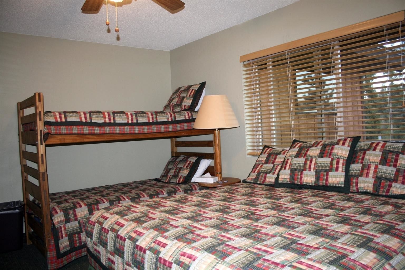 Quarto com beliche, cobertor estampado xadrez, escada de madeira, abajur e janela com persianas.