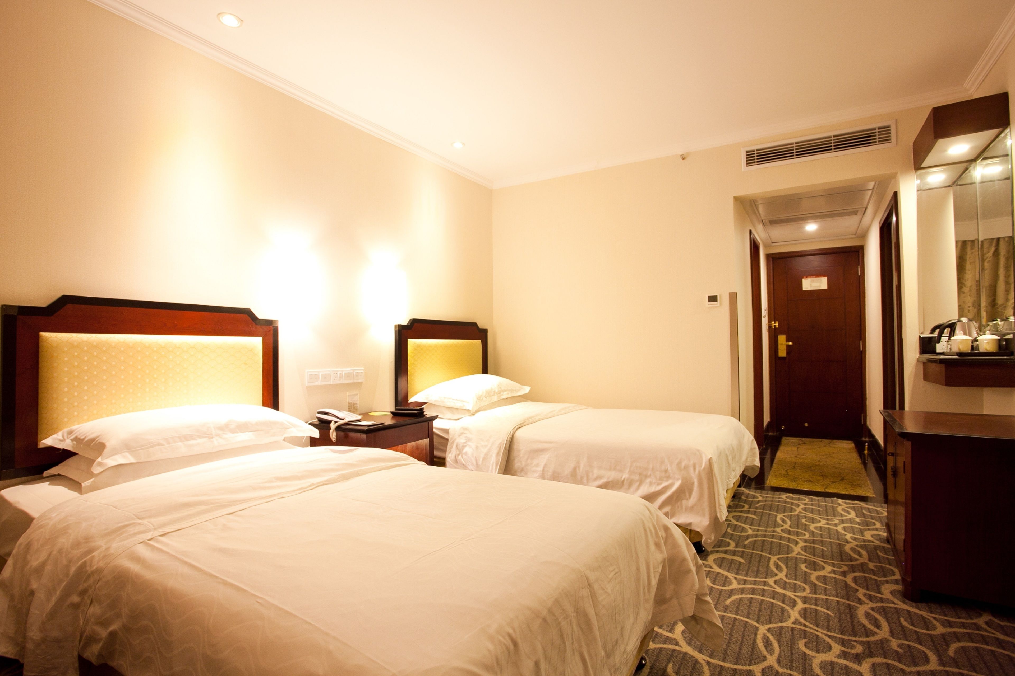 Quarto de hotel com duas camas, mesa de cabeceira, escrivaninha com abajur, espelho e porta que dá para um corredor.