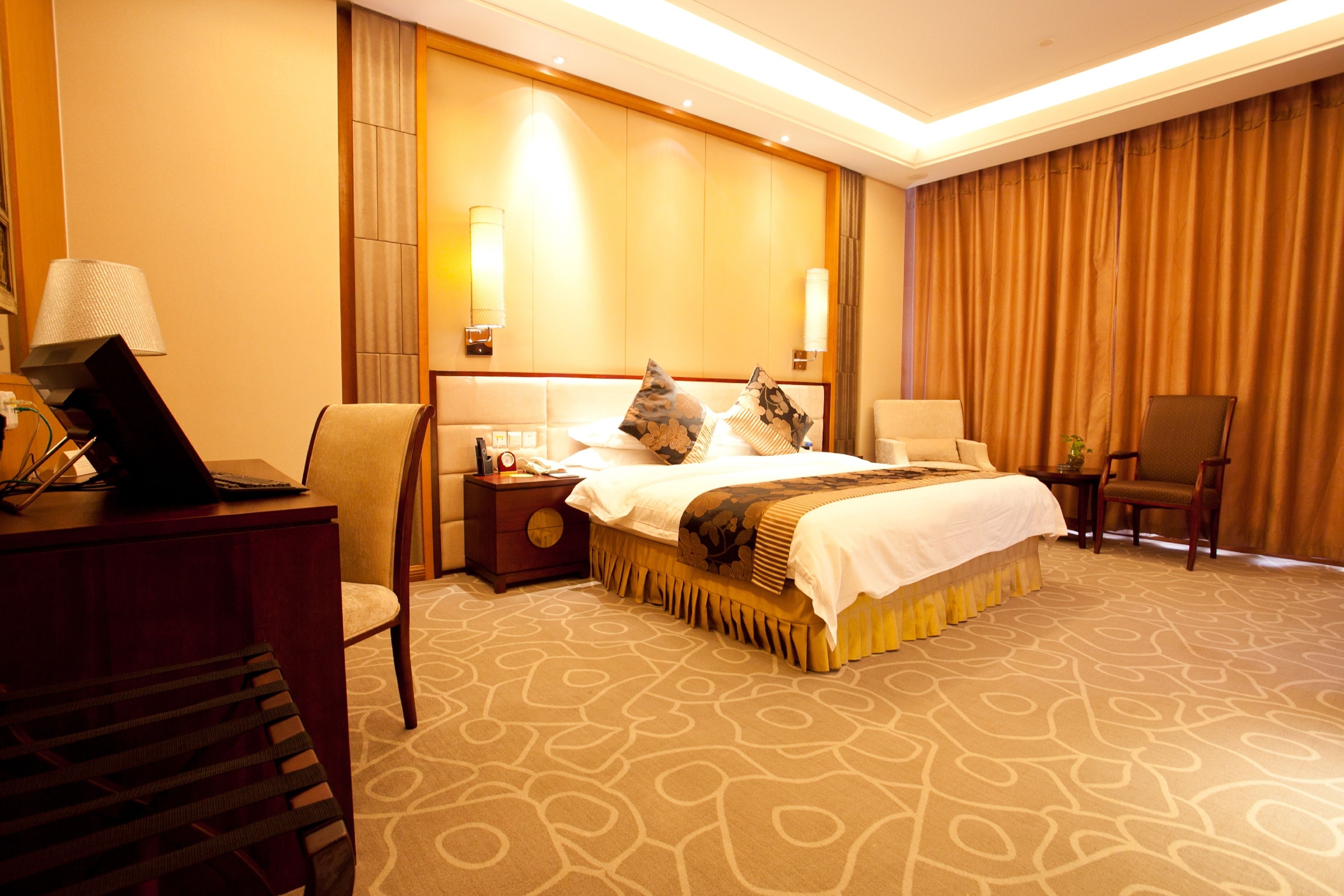 Quarto de hotel com uma cama grande, uma escrivaninha com um laptop, uma cadeira, uma mesa de cabeceira e um abajur.