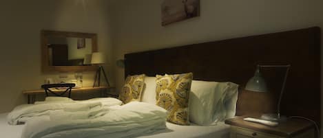 Cama bem arrumada com lençóis brancos, mesa de cabeceira de madeira com abajur, e escrivaninha com abajur e espelho.