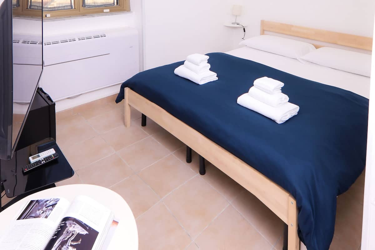 Quarto de hotel com cama, televisão, pintura e janela com vista para edifícios.