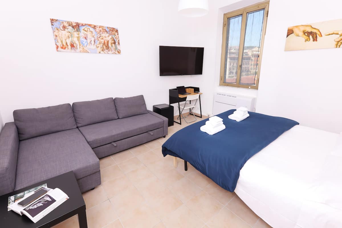 Quarto de hotel moderno com um sofá cinzento, uma cama com cobertura azul, uma televisão de tela plana, uma pequena mesa com um laptop e uma janela com vista para a cidade.
