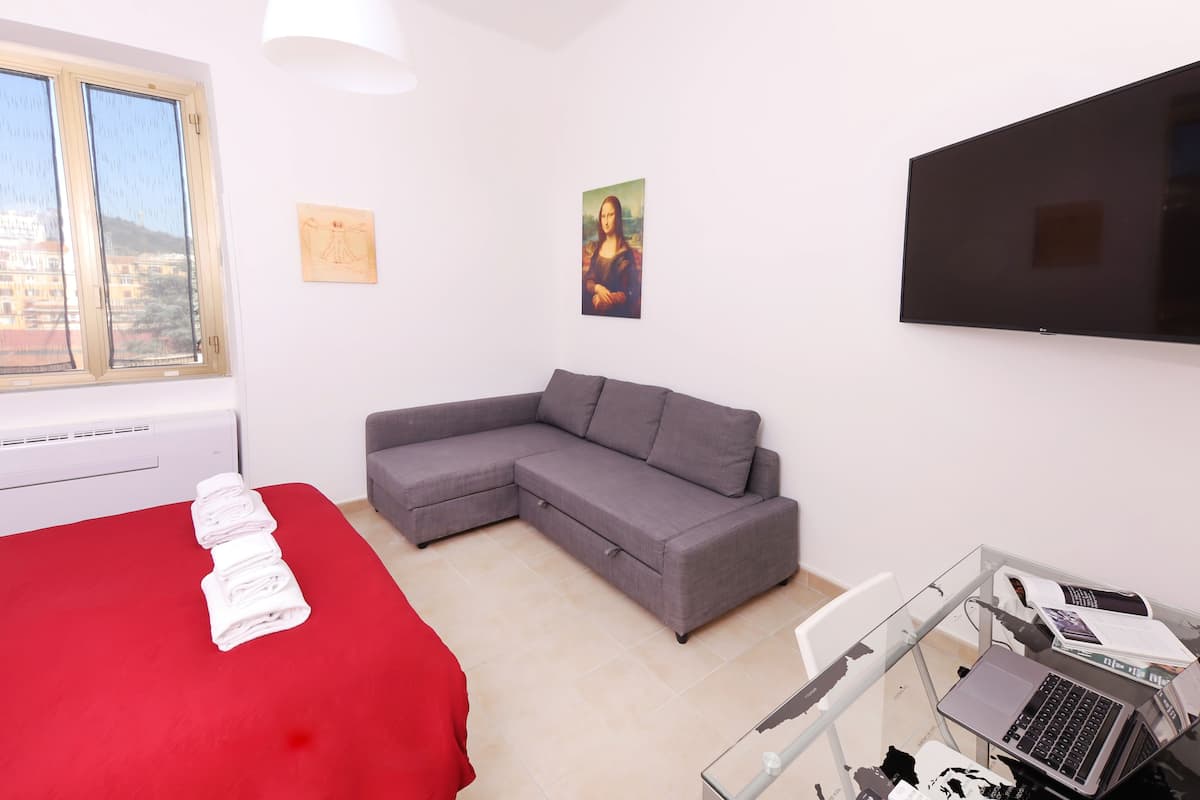 Quarto moderno com uma cama, sofá cinzento, televisão e escrivaninha com um computador portátil.