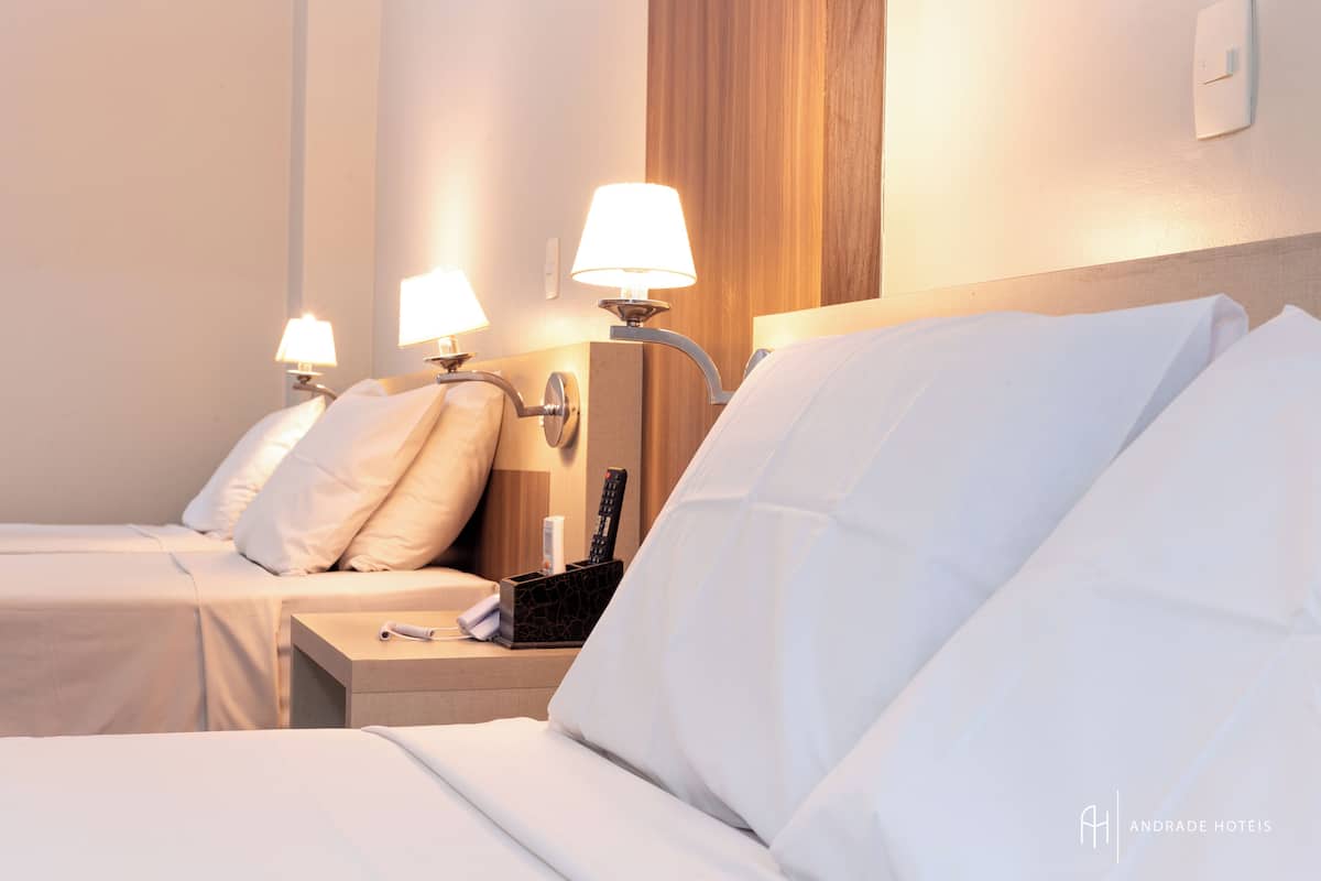 Quarto de hotel com duas camas, cada uma com roupa de cama e almofadas brancas, uma mesa de cabeceira com luminária e suporte para controle remoto.