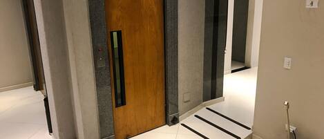 Um corredor com porta de madeira identificada pela letra 'T' para o banheiro, corrimão fixado na parede e um pequeno degrau que leva a outro cômodo.
