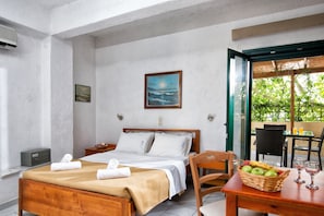 Quarto com cama, mesas de cabeceira, uma cadeira de madeira, uma mesa de jantar com frutas e copos, e um quadro na parede.