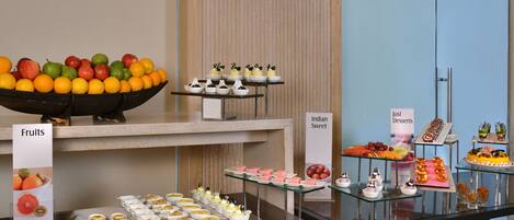 Buffet com diversas opções de comida, incluindo frutas, doces indianos e sobremesas.