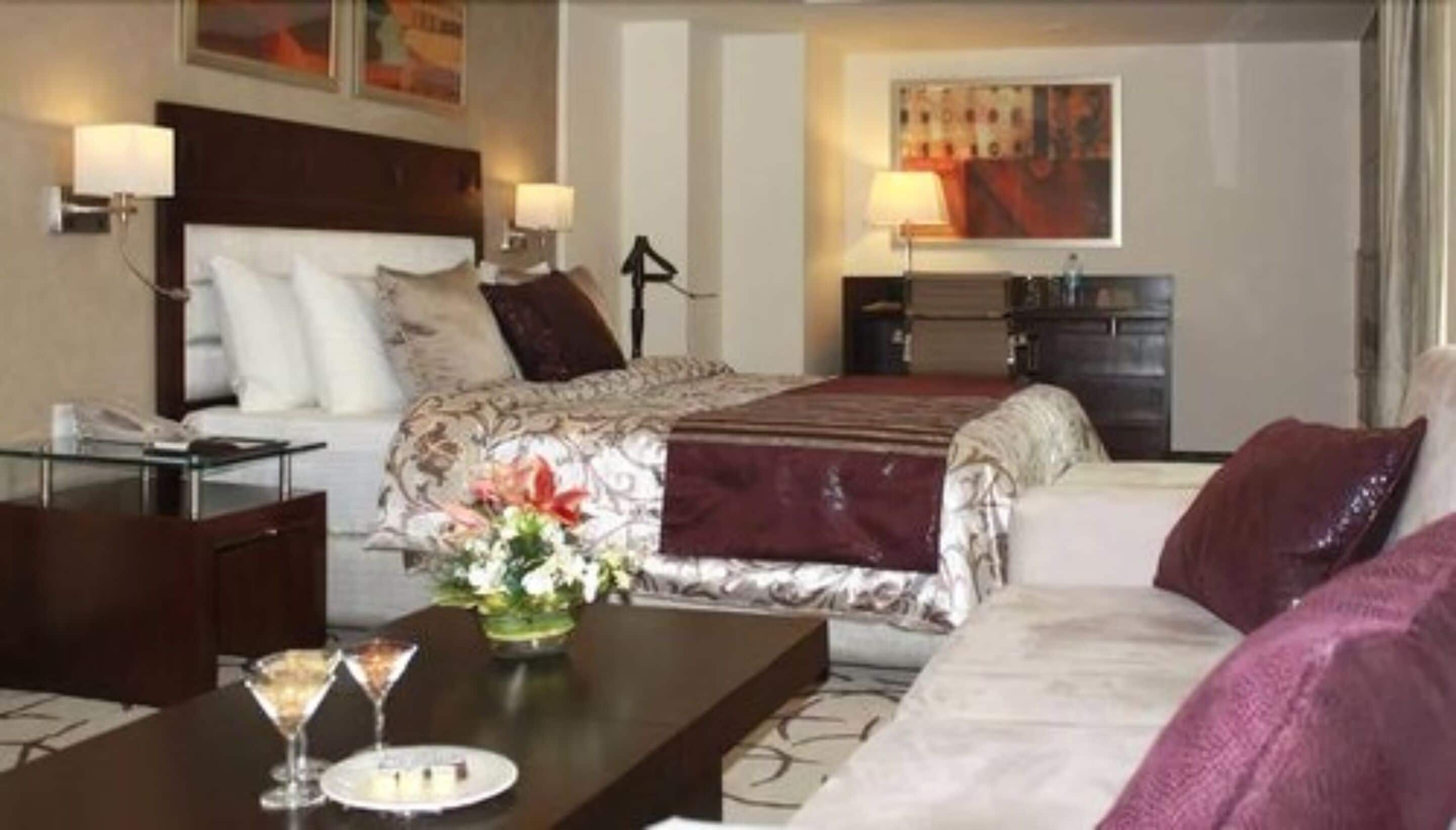Quarto de hotel com cama, sofá, escrivaninha e mesa de jantar, com um vaso de flores e uma taça de vinho.