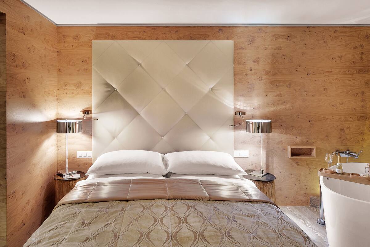 Quarto com cabeceira estofada, cama com roupa de cama estampada, abajures e painéis de madeira na parede.