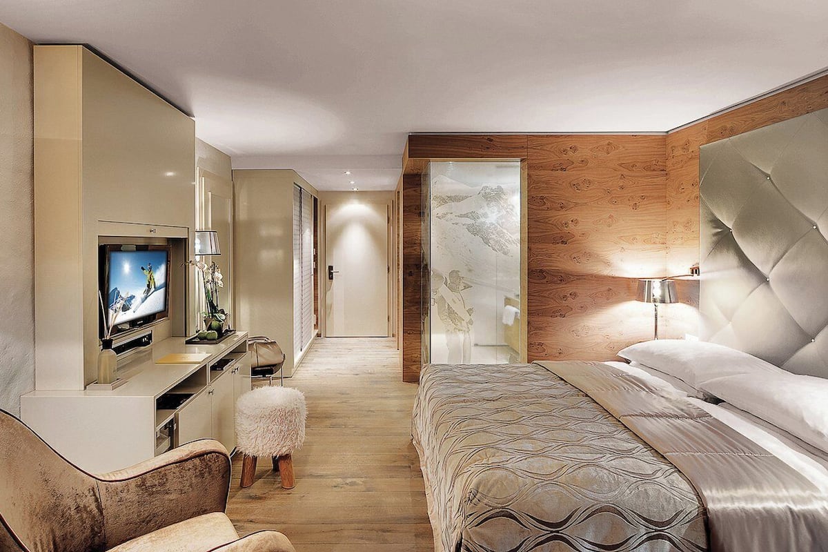 Quarto de hotel moderno com uma cama grande, uma escrivaninha com televisão de tela plana, um espaço de estar com uma poltrona e um corredor que leva a outro quarto.