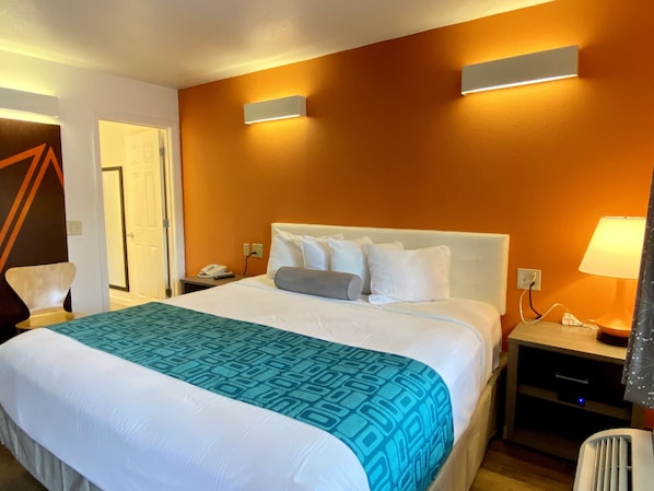 Quarto de hotel com uma cama grande, uma mesinha de cabeceira, uma luminária e um ponto de luz na parede.