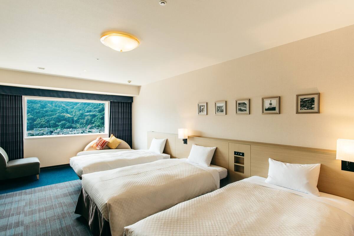 Quarto de hotel com três camas, uma escrivaninha, uma cadeira e uma janela grande com vista para a cidade.