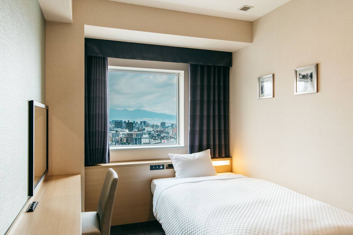 Quarto de hotel com cama, escrivaninha, cadeira, janela com vista para a cidade e duas imagens emolduradas na parede.