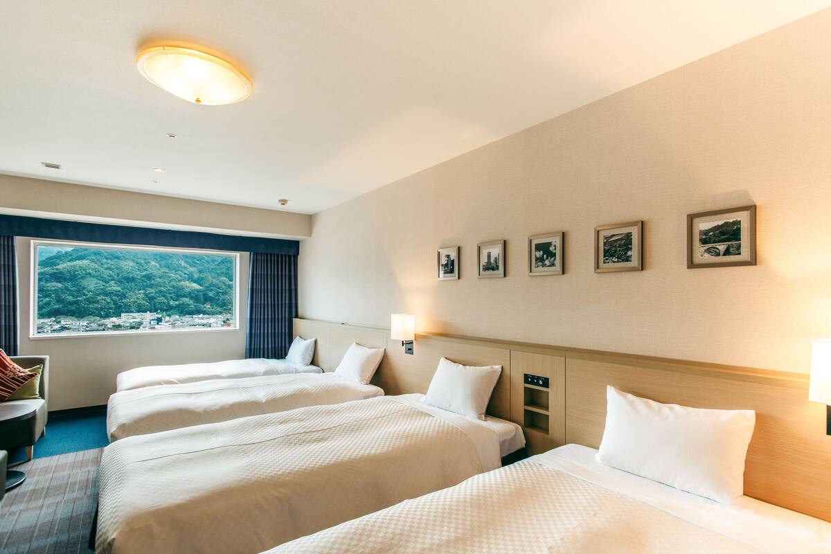 Quarto de hotel com três camas, uma janela ampla com vista para a cidade e parede com quadros emoldurados.