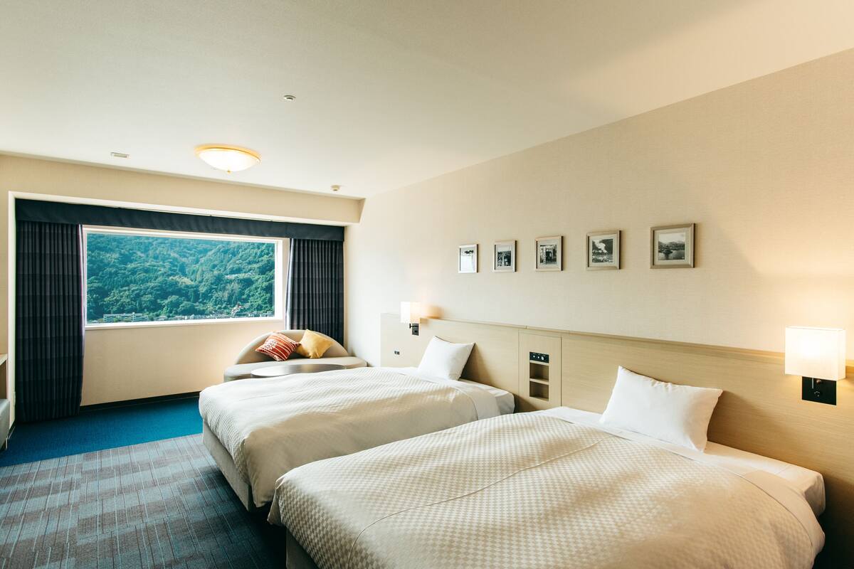Quarto de hotel com duas camas, uma janela ampla com vista para uma floresta e uma parede com quadros.