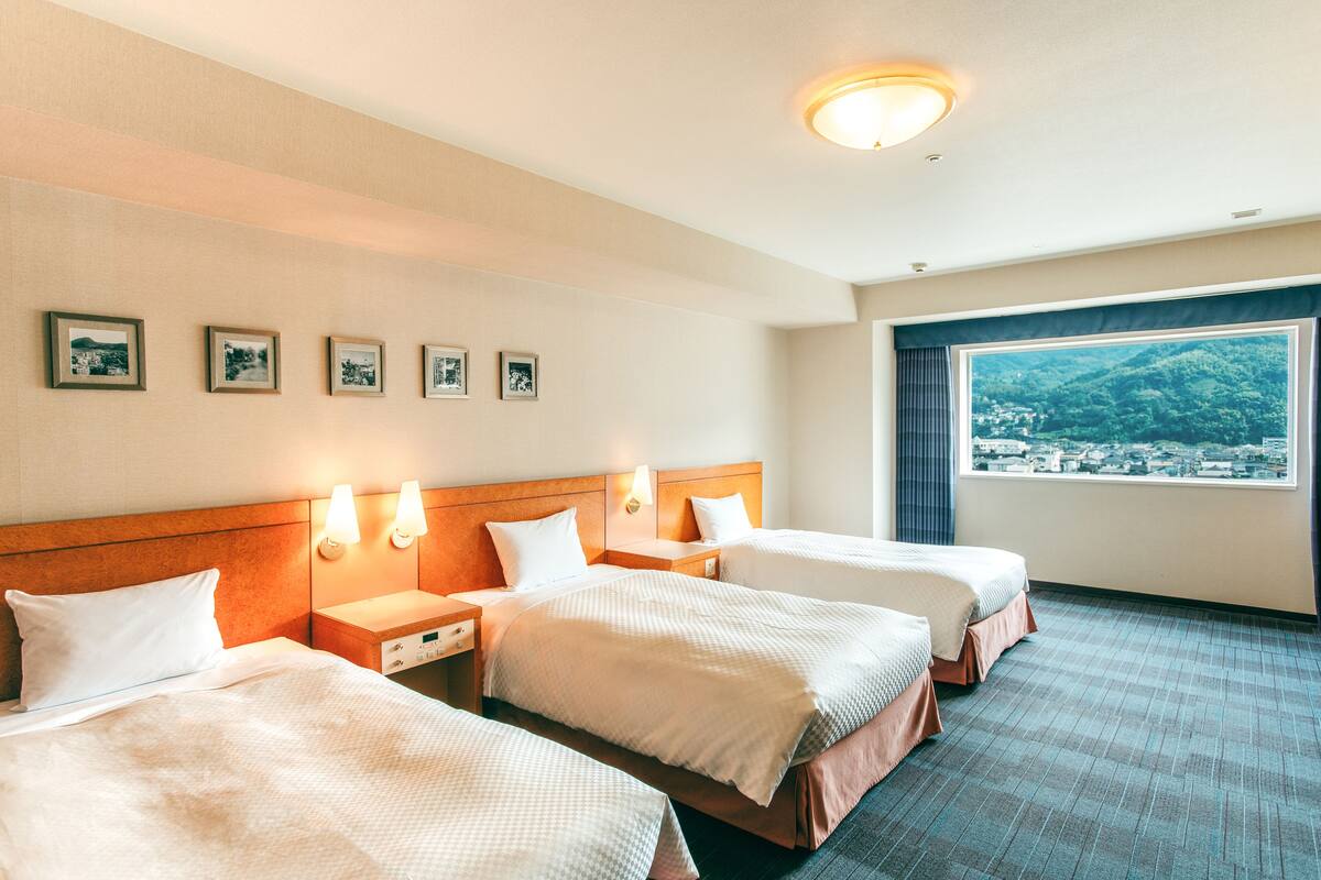 Quarto de hotel com três camas, uma janela grande com vista para a cidade e luminárias fixadas na parede.
