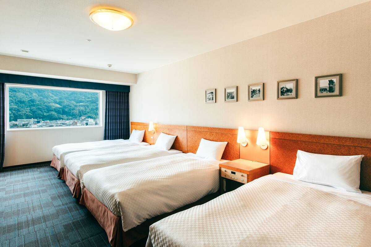Quarto de hotel com três camas, uma janela grande com vista para a cidade e várias fotos emolduradas na parede.