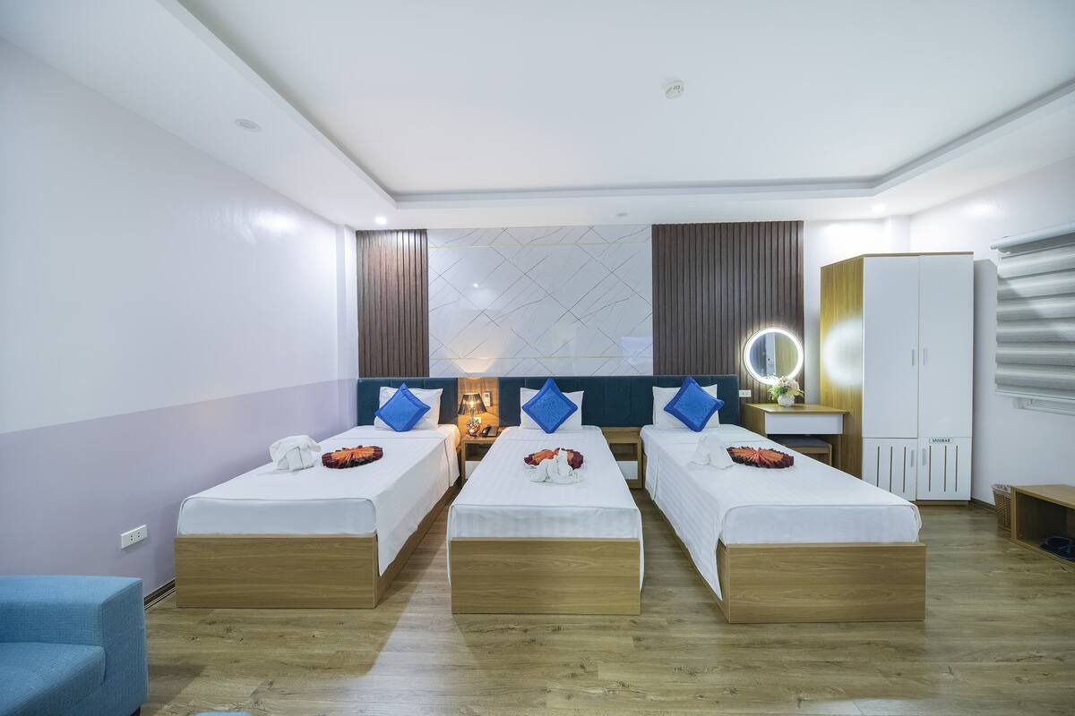 Quarto de hotel com três camas de solteiro, um guarda-roupa de madeira, uma escrivaninha com espelho e uma poltrona azul.
