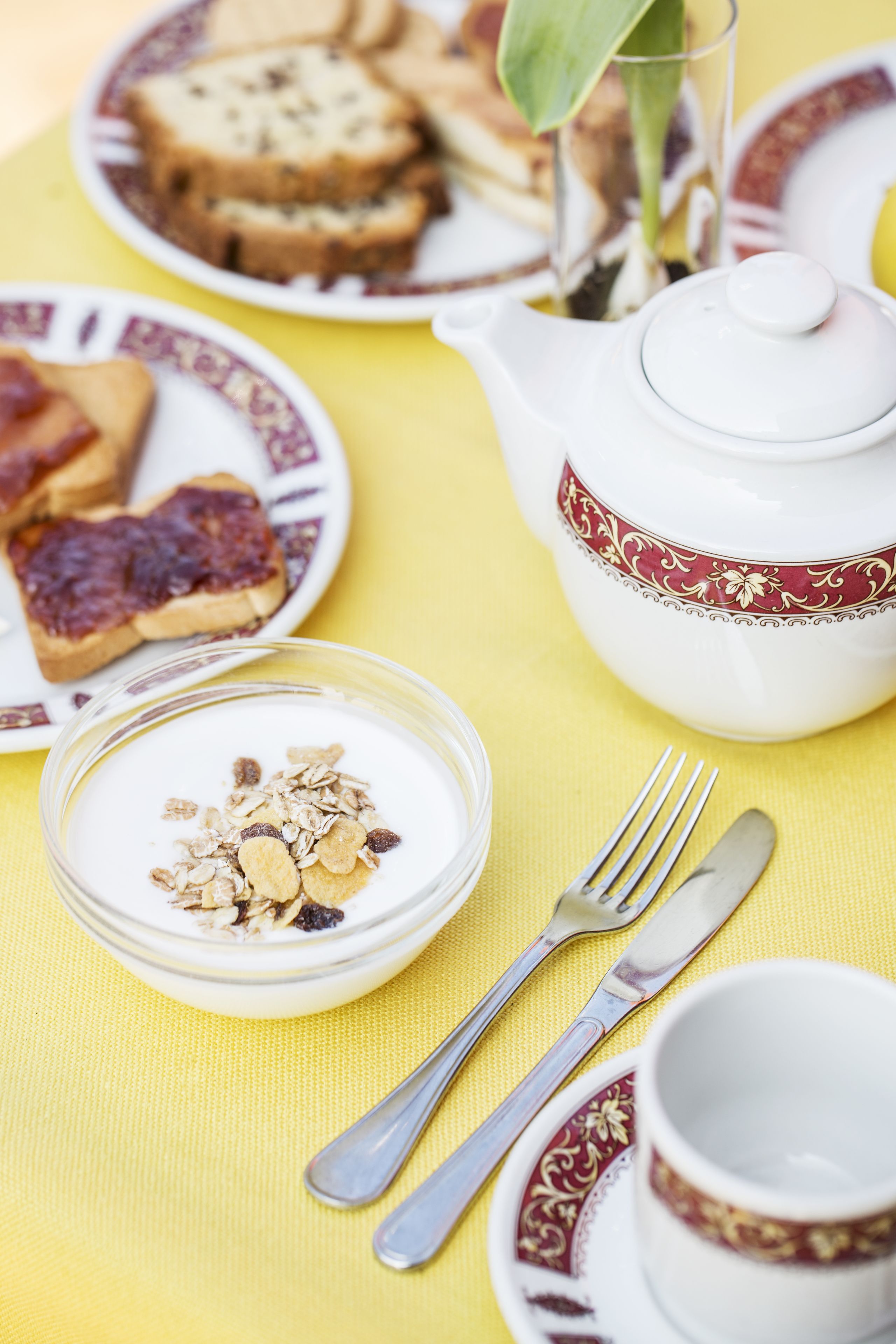 Uma mesa posta com um bule, uma tigela de iogurte com granola, uma xícara, um prato com torradas e talheres.