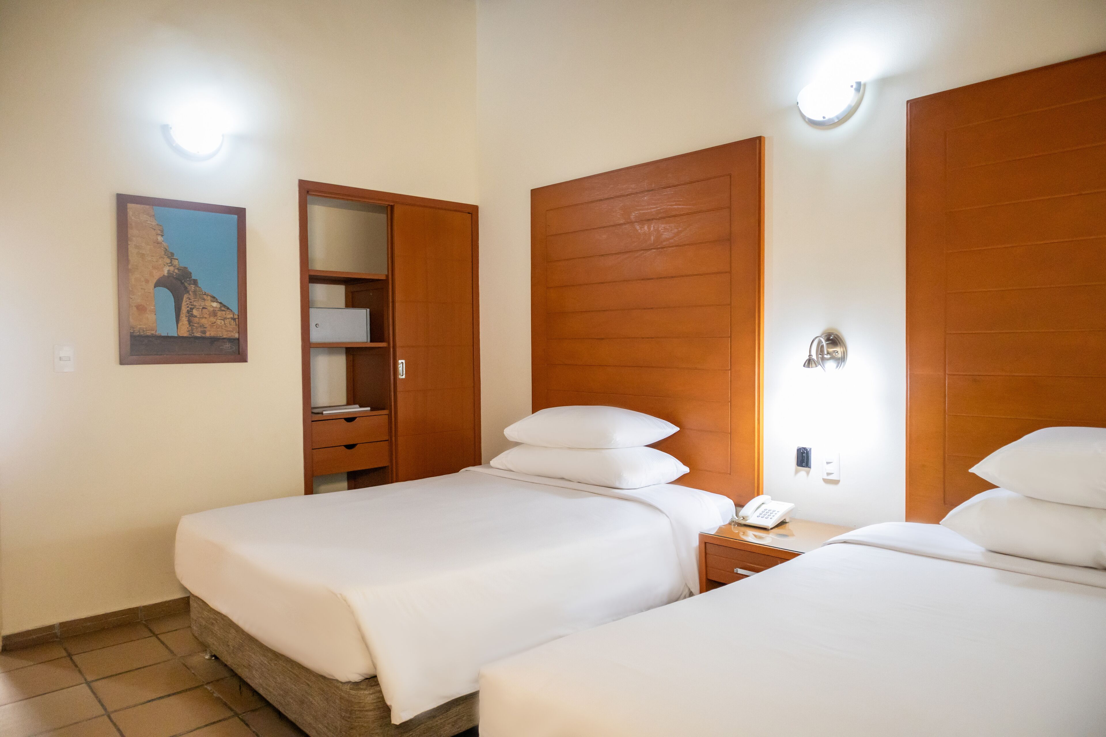 Quarto de hotel com duas camas, uma cabeceira de madeira, uma mesinha de cabeceira com telefone e um pequeno armário.