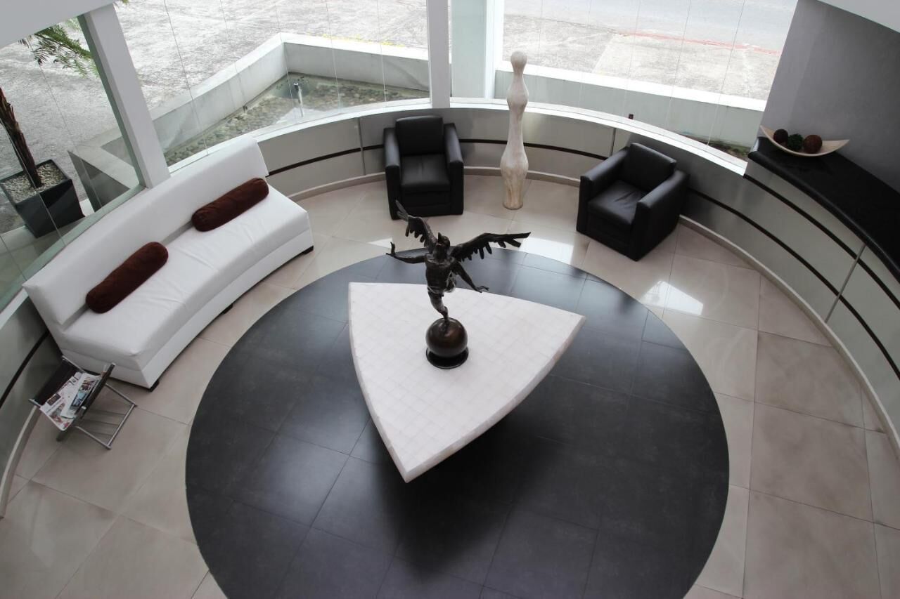 Um lobby moderno com uma peça escultórica branca central, assentos de couro preto e um guarda-corpo de vidro com vista para um pátio.