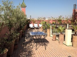 Terraço no telhado com mesa e cadeiras de metal, rodeado por plantas em vasos e piso de telhas vermelhas.