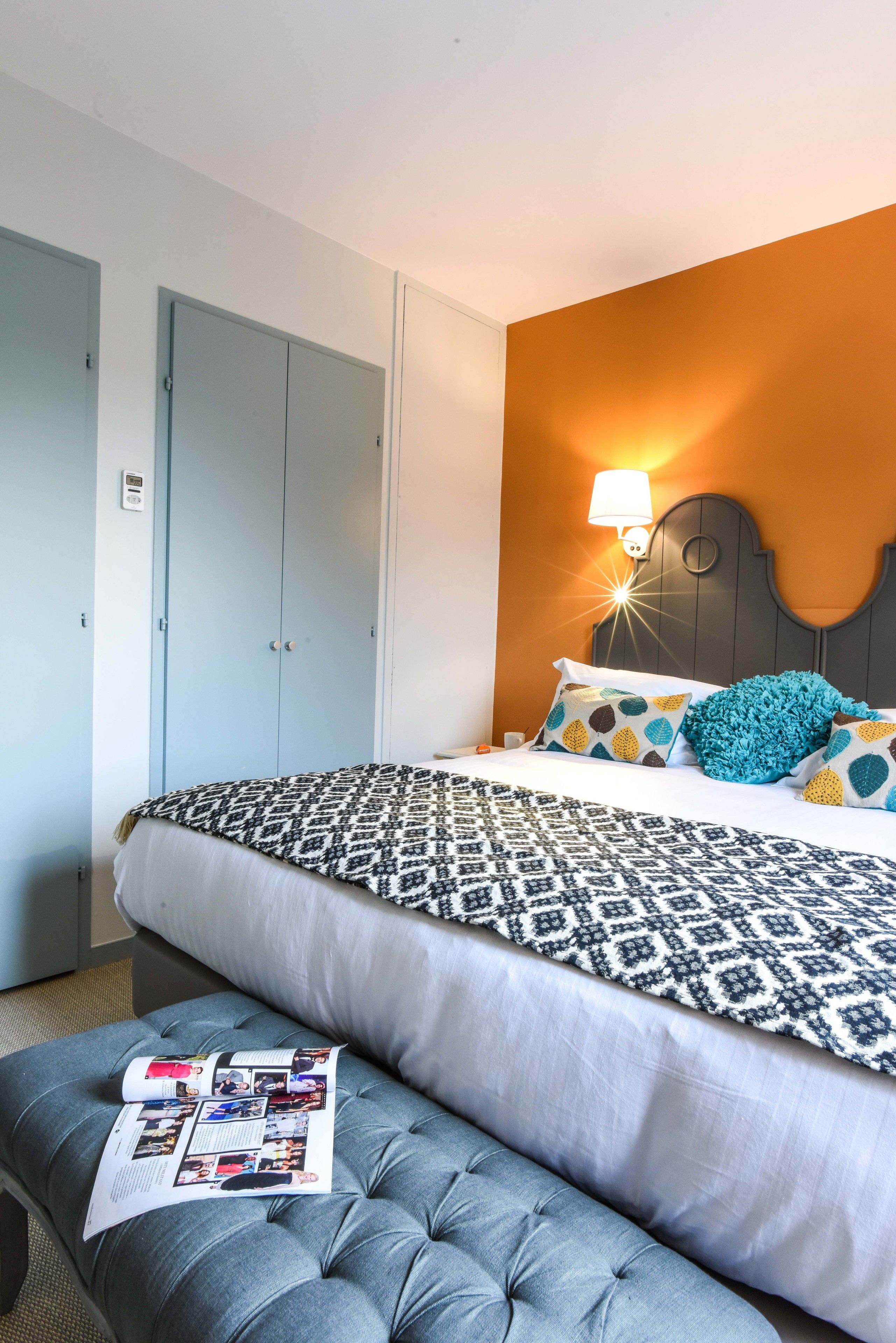 Quarto de hotel com uma cama, uma luminária de cabeceira e uma revista no banquinho.